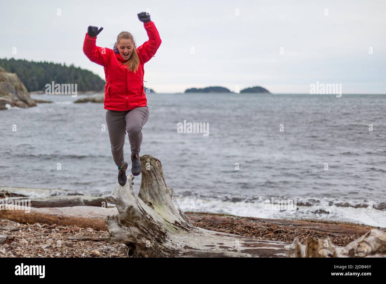 Woman in a red jack leaps in the air from driftwood on the beach along ...