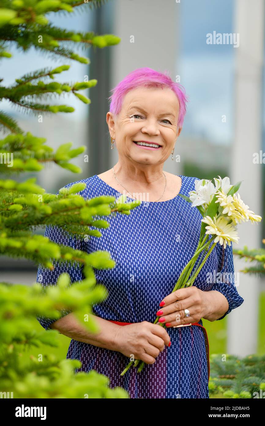 Portrait of smiling mature woman with short pink hair standing near fir ...