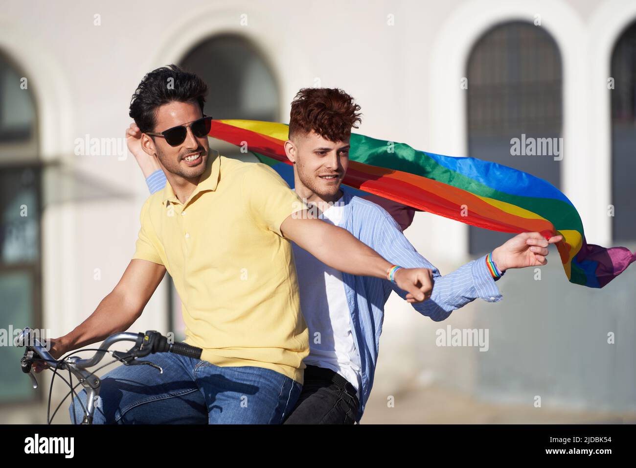 Young couple of friends holding a gay pride flag while riding a bike ...