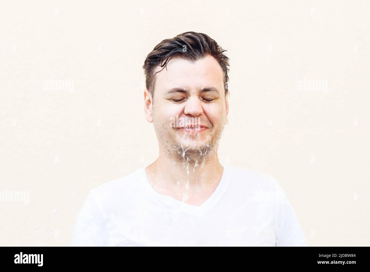 Wet young caucasian man in white t shirt with closed eyes and water is ...