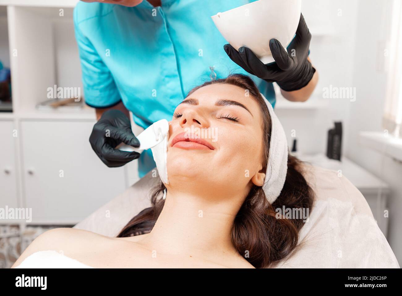 Facial beauty procedure. Beautician in latex gloves applying with spatula rejuvenation mask on ...