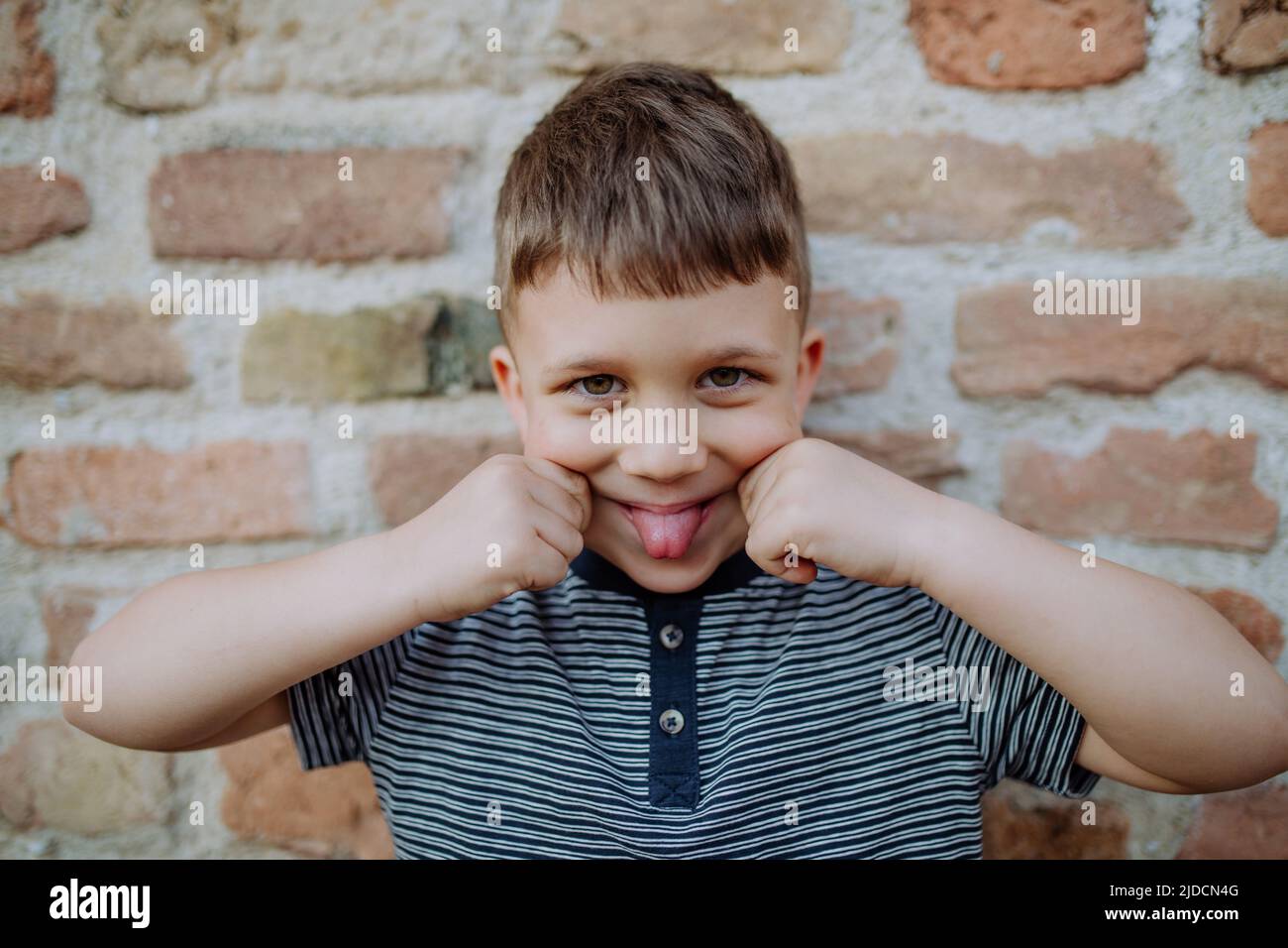 Little boy standing by brick wall and making funny faces in street ...
