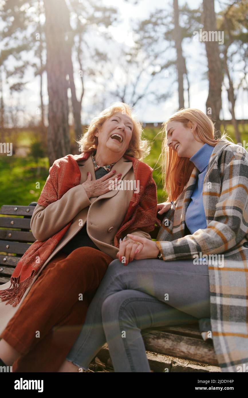 A young ginger woman is sitting with her grandmother and talking on the park bench in the cold ...