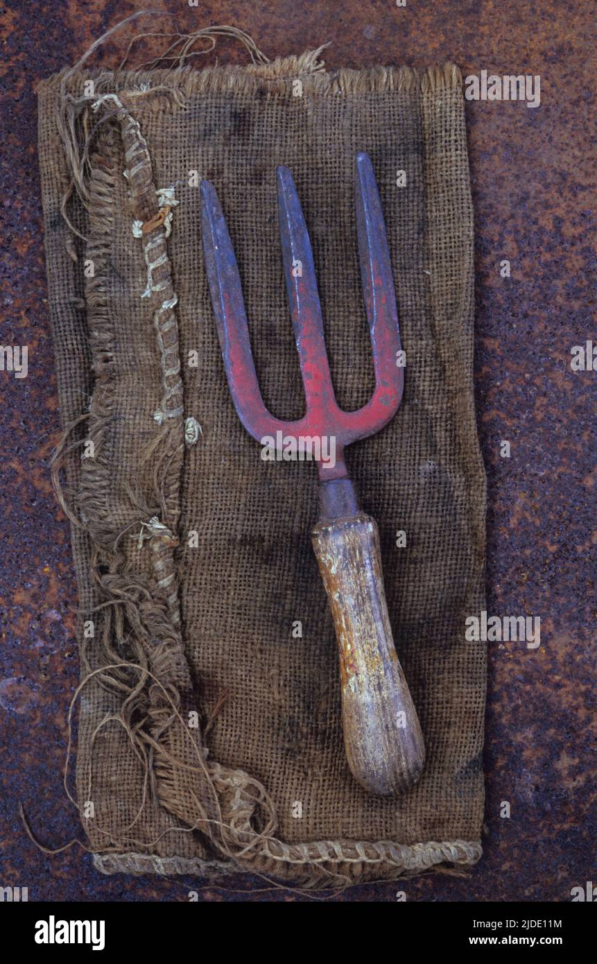 Well-used garden hand fork lying on hessian sack on rusty metal sheet ...
