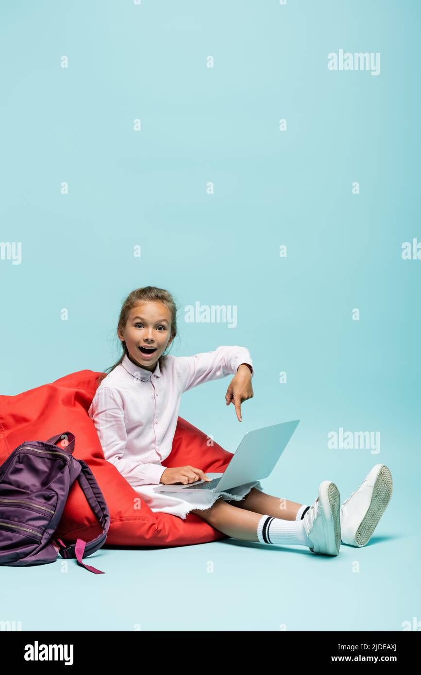 Positive schoolkid pointing with finger at laptop near backpack on blue ...