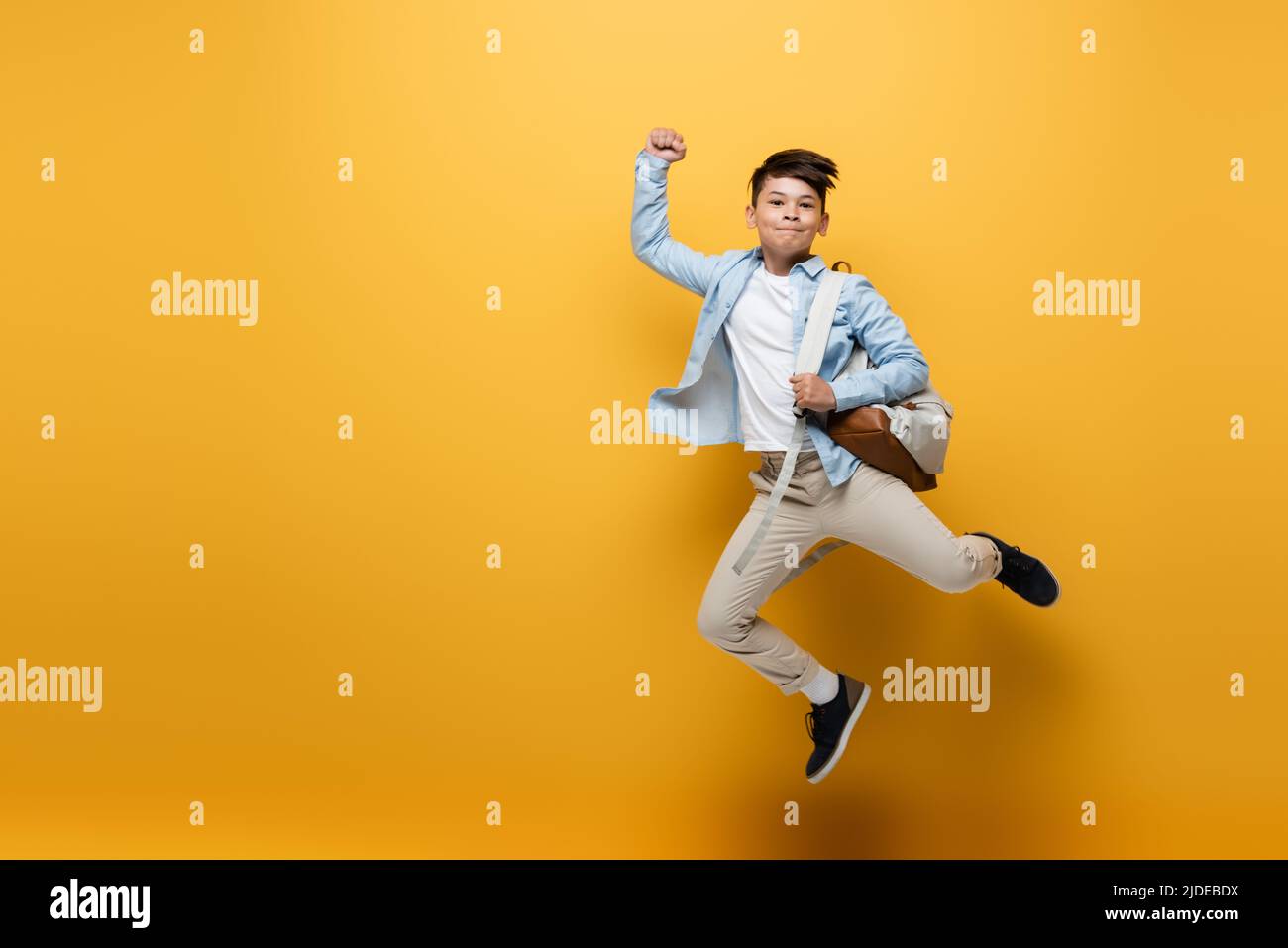 Asian schoolkid holding backpack and showing yes gesture while jumping ...
