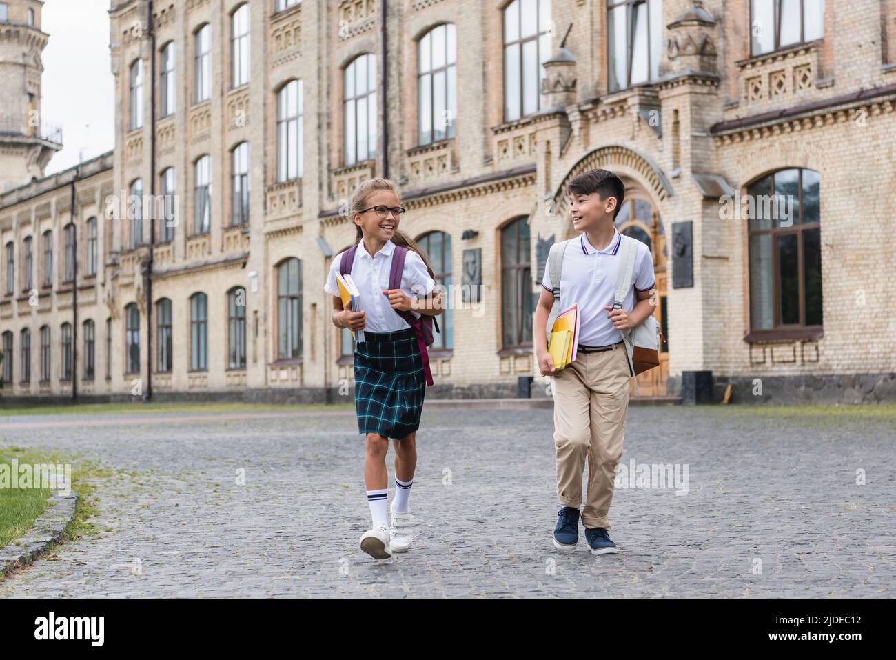 Schoolgirl with notebooks talking to asian friend with backpack while ...