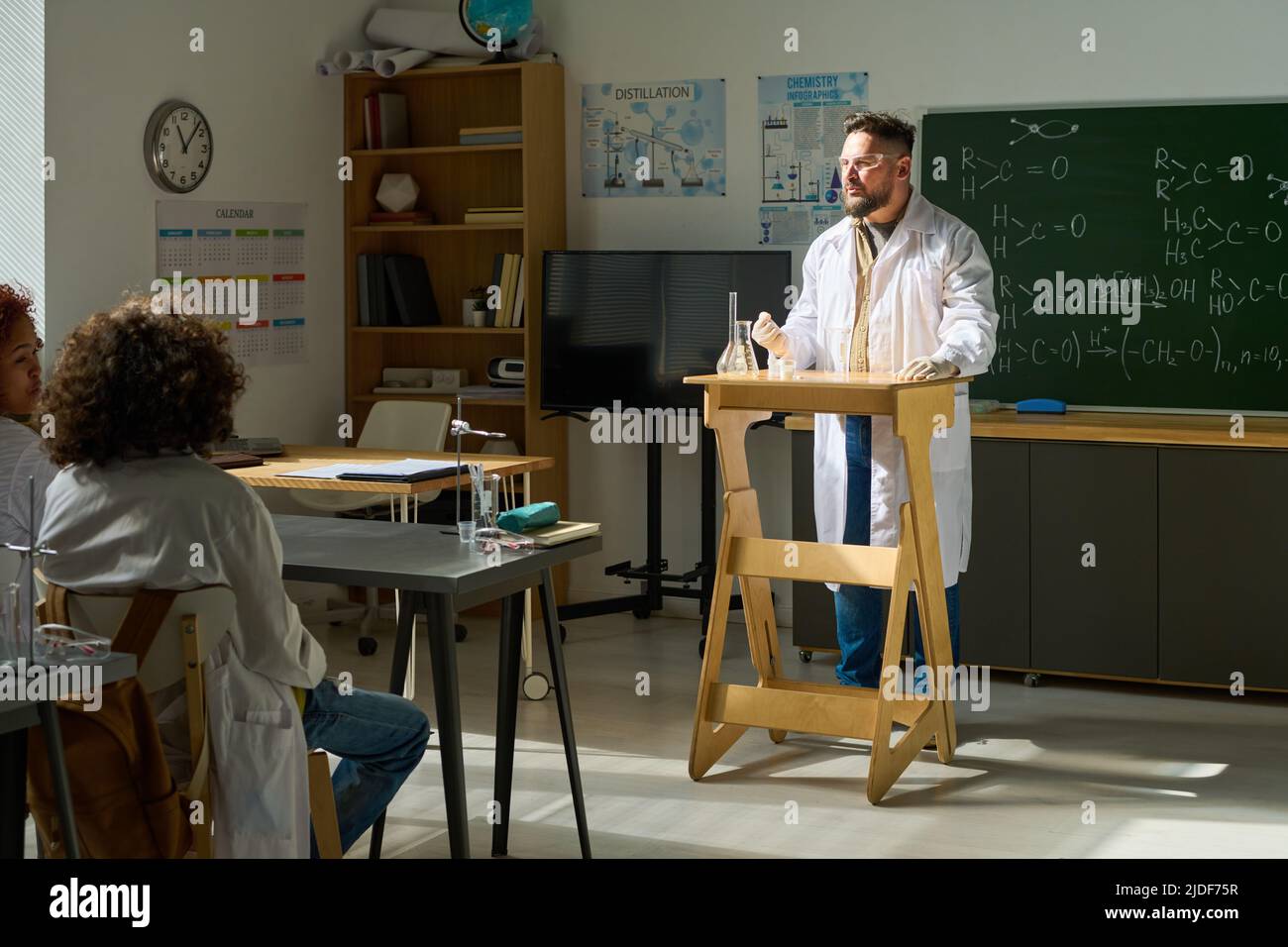 Confident teacher of chemistry in labcoat standing by desk in front of ...