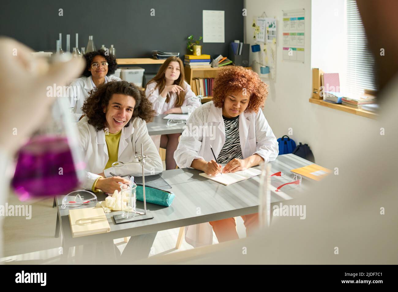 Happy teenage guy in labcoat looking at teacher of chemistry showing ...