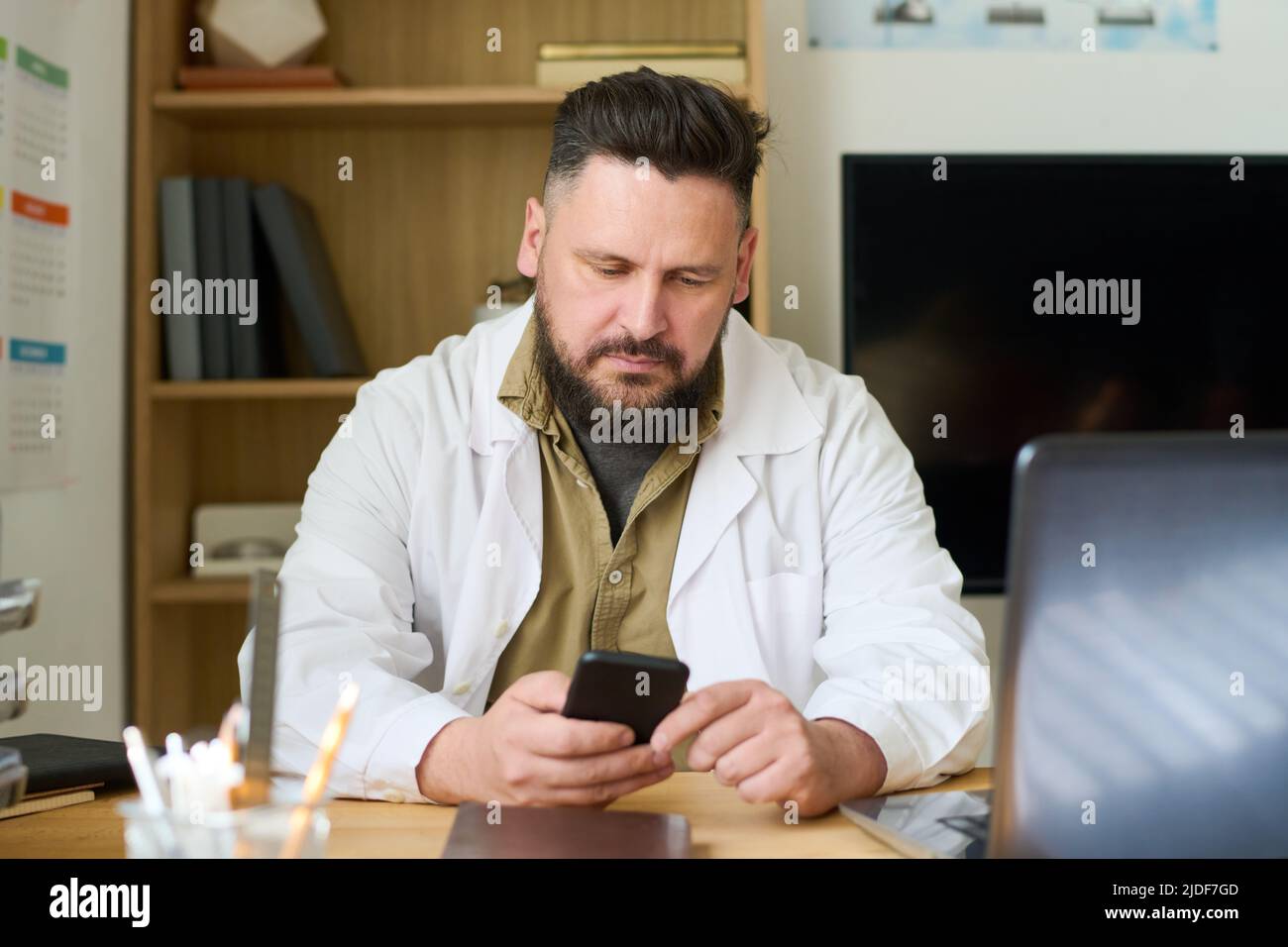 Serious mature teacher in labcoat scrolling in smartphone or texting ...