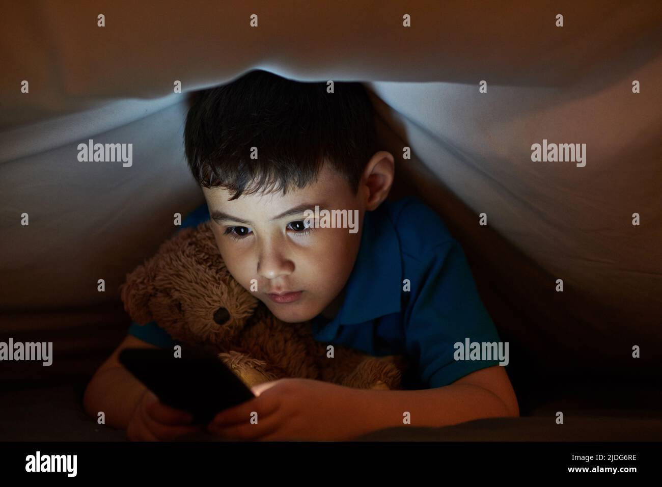 Kid hugging teddy bear when playing game on smartphone under blanket at ...