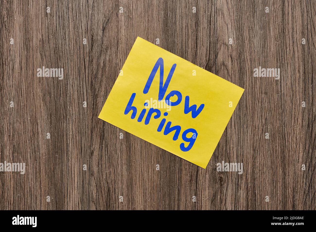 Now hiring text on yellow note with blue sheet on wooden desk. Consept ...