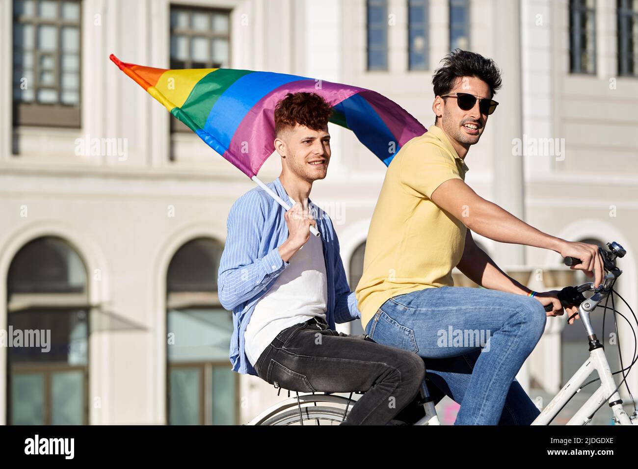 Young Gay Couple Holding A Gay Pride Flag While Riding A Bike Outdoors Lgbt Relationship And