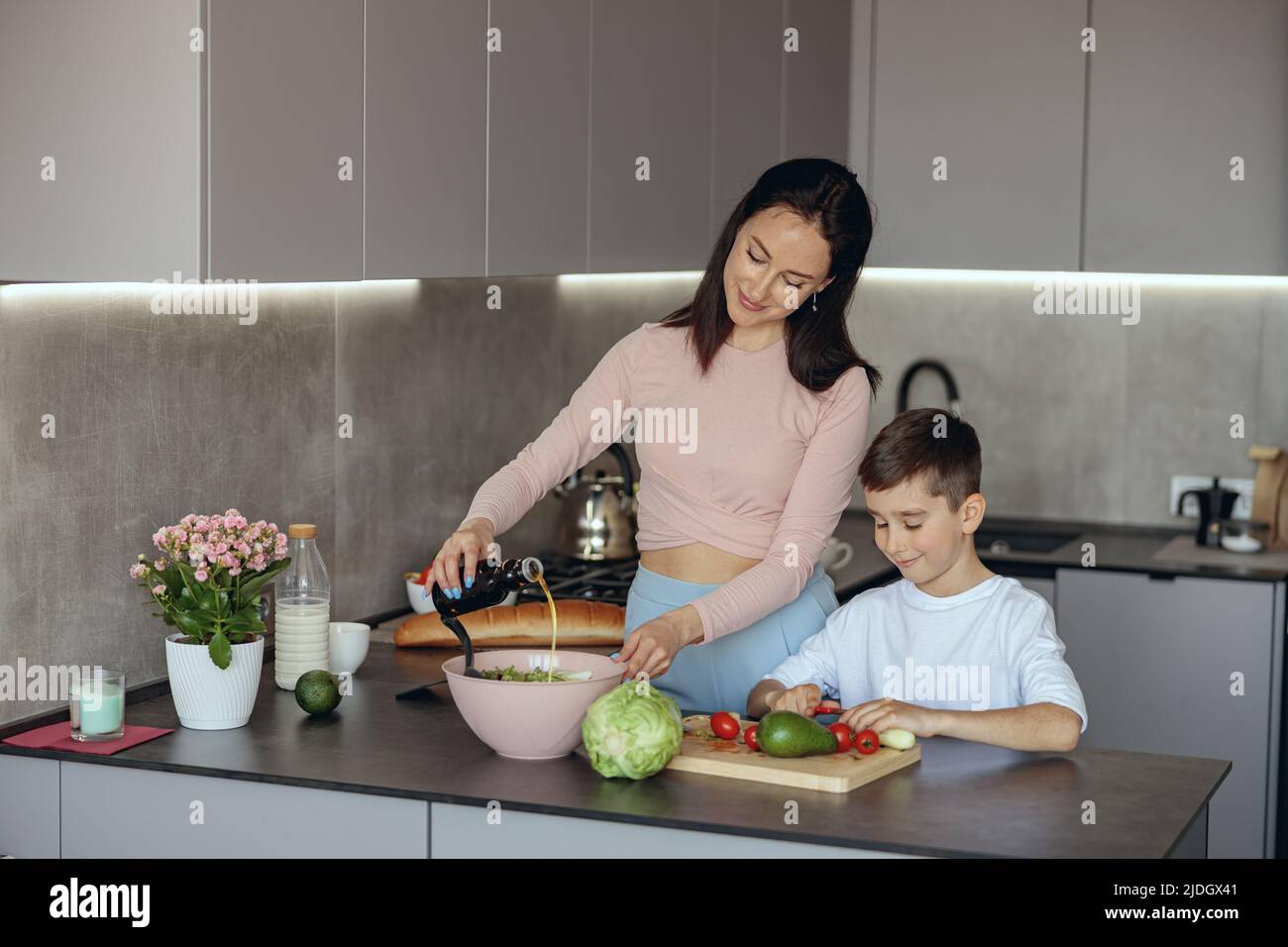 Pretty young mother cooking salat and putting olive oil in, small son ...