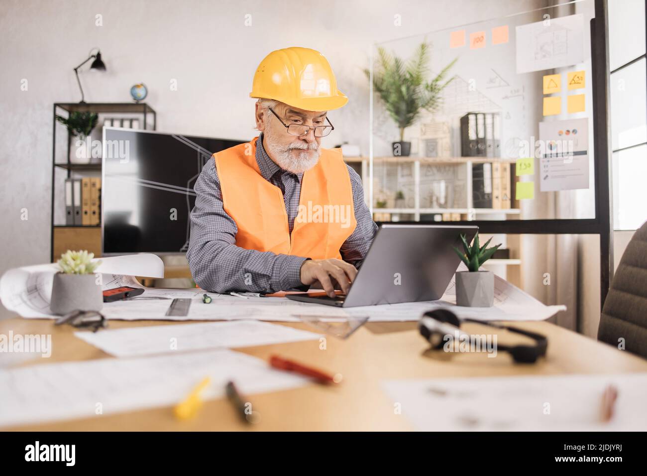 Bearded engineer senior man in helmet and reflective vest sitting at ...