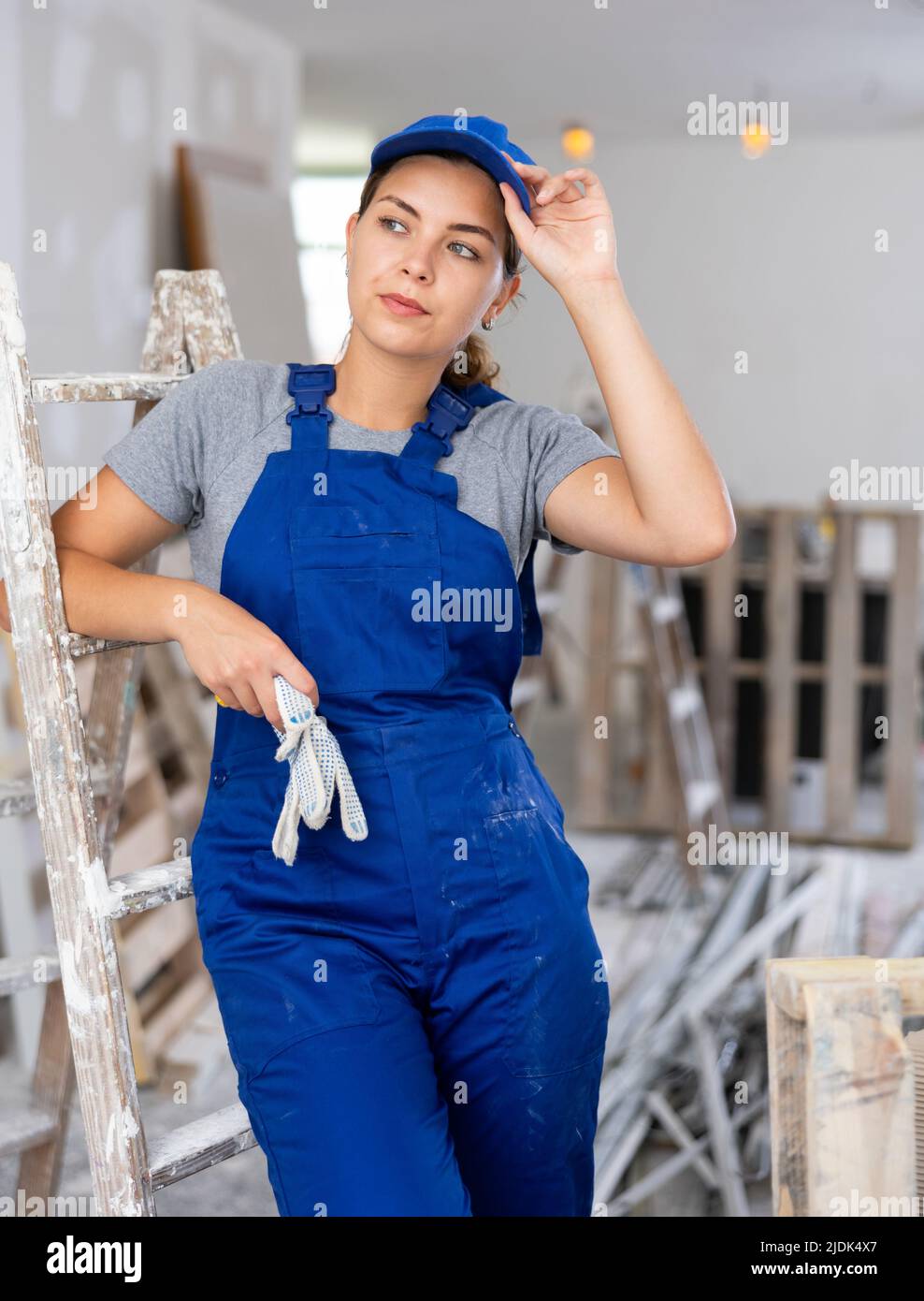 Portrait of positive builder woman in blue overalls next to stepladder ...