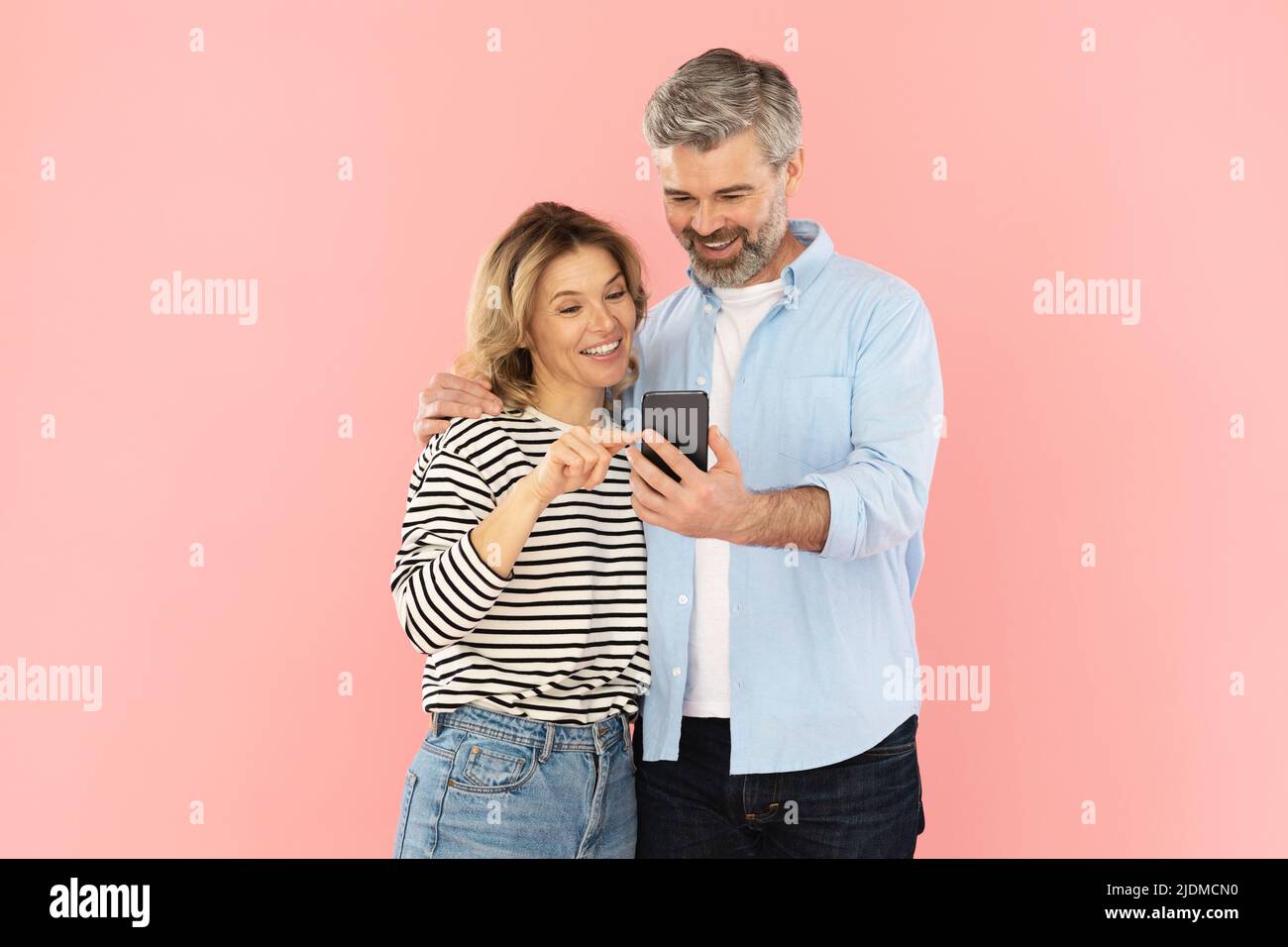 Cheerful Middle Aged Spouses Using Mobile Phone On Pink Background ...