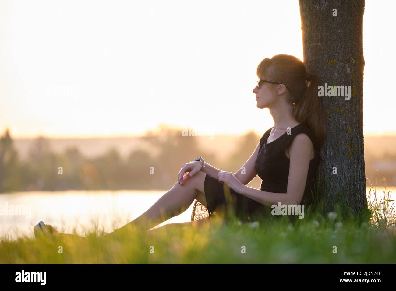 Lonely woman sitting alone on green grass lawn leaning to tree trunk on ...