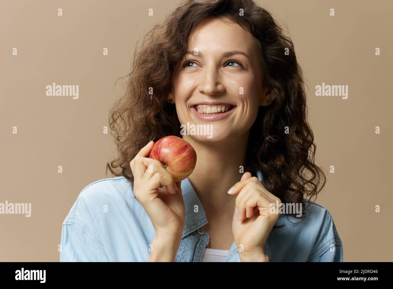 Cheerful curly cute lady in jeans casual shirt with apple near cheek ...