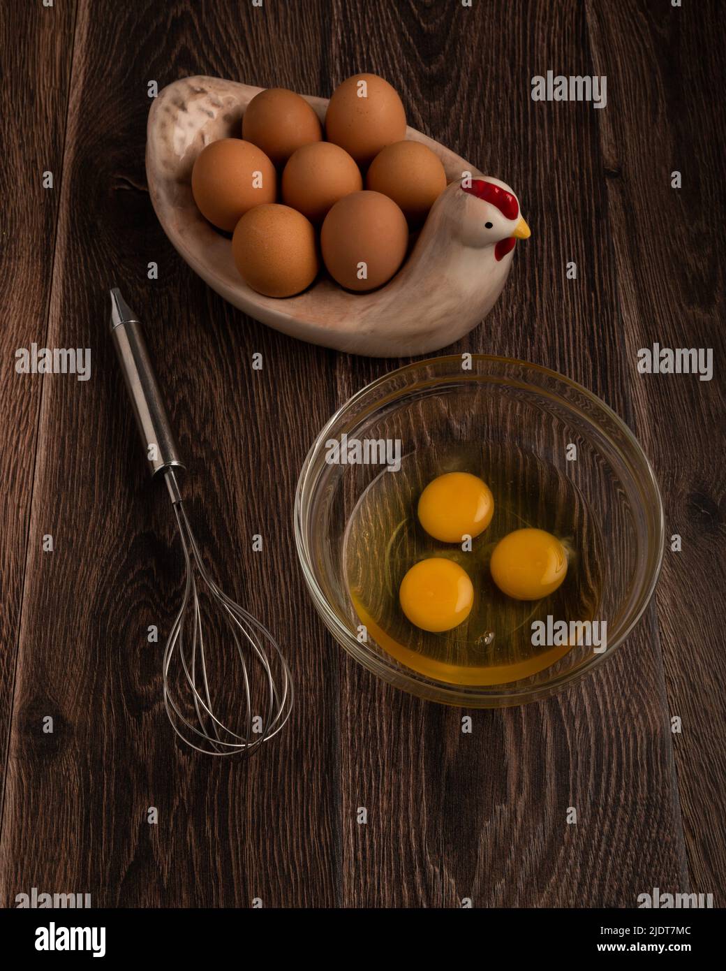 A whisk, chicken eggs in a glass bowl and in a chicken shaped dish on a ...