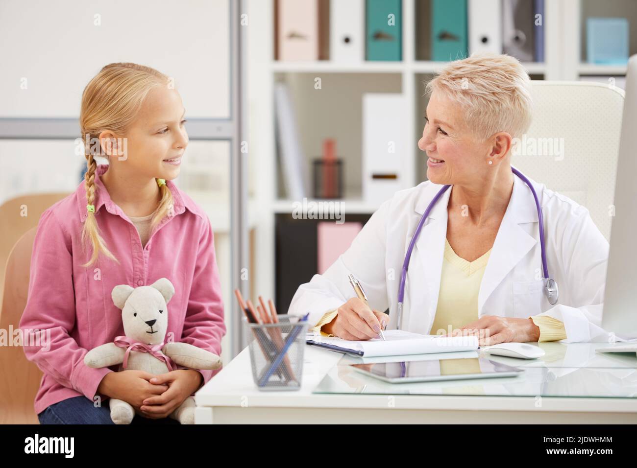 Cheerful pretty girl sitting on chair by doctors table and holding toy ...