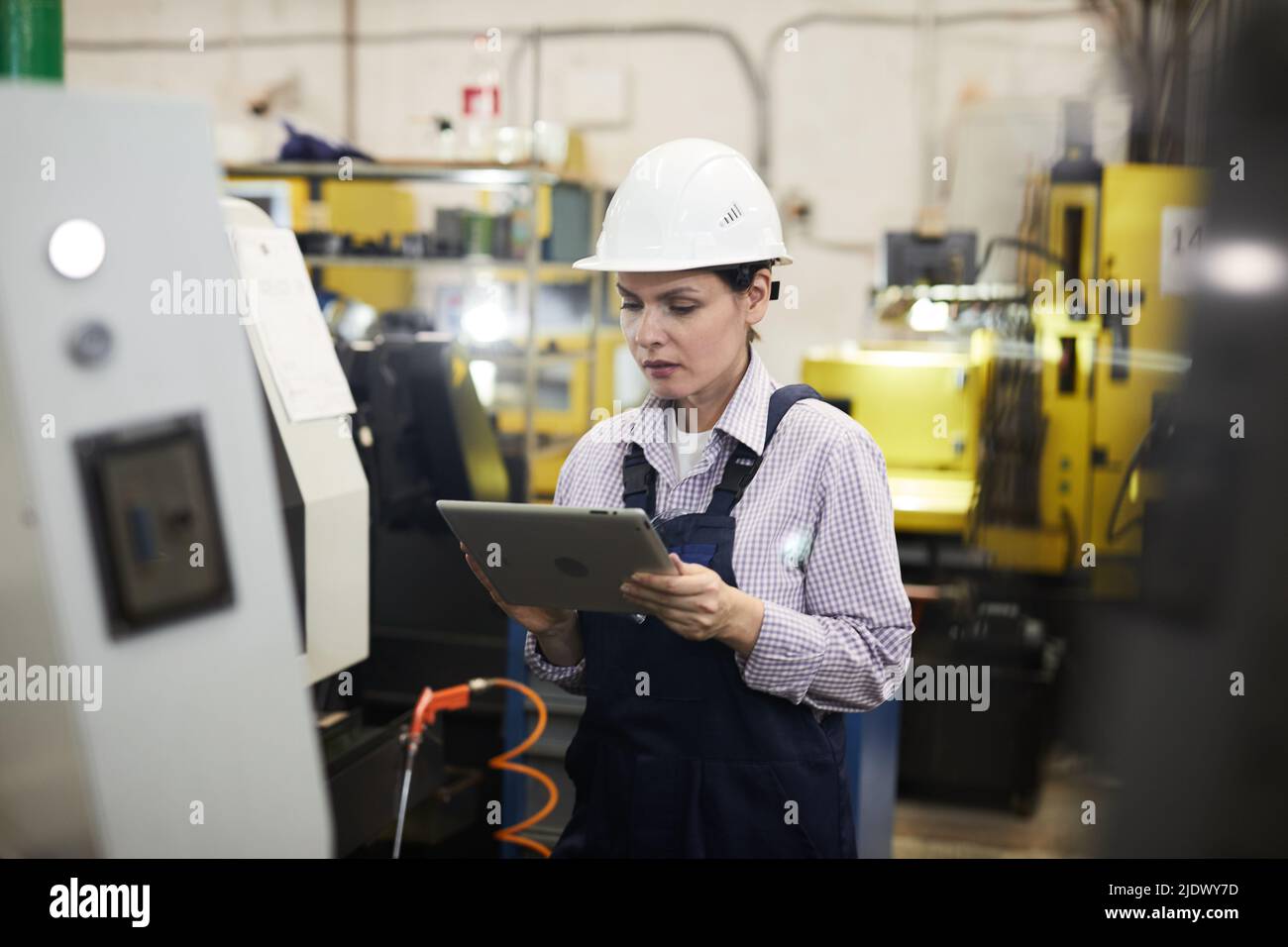 Concentrated young female cnc machine operator in hardhat standing at ...