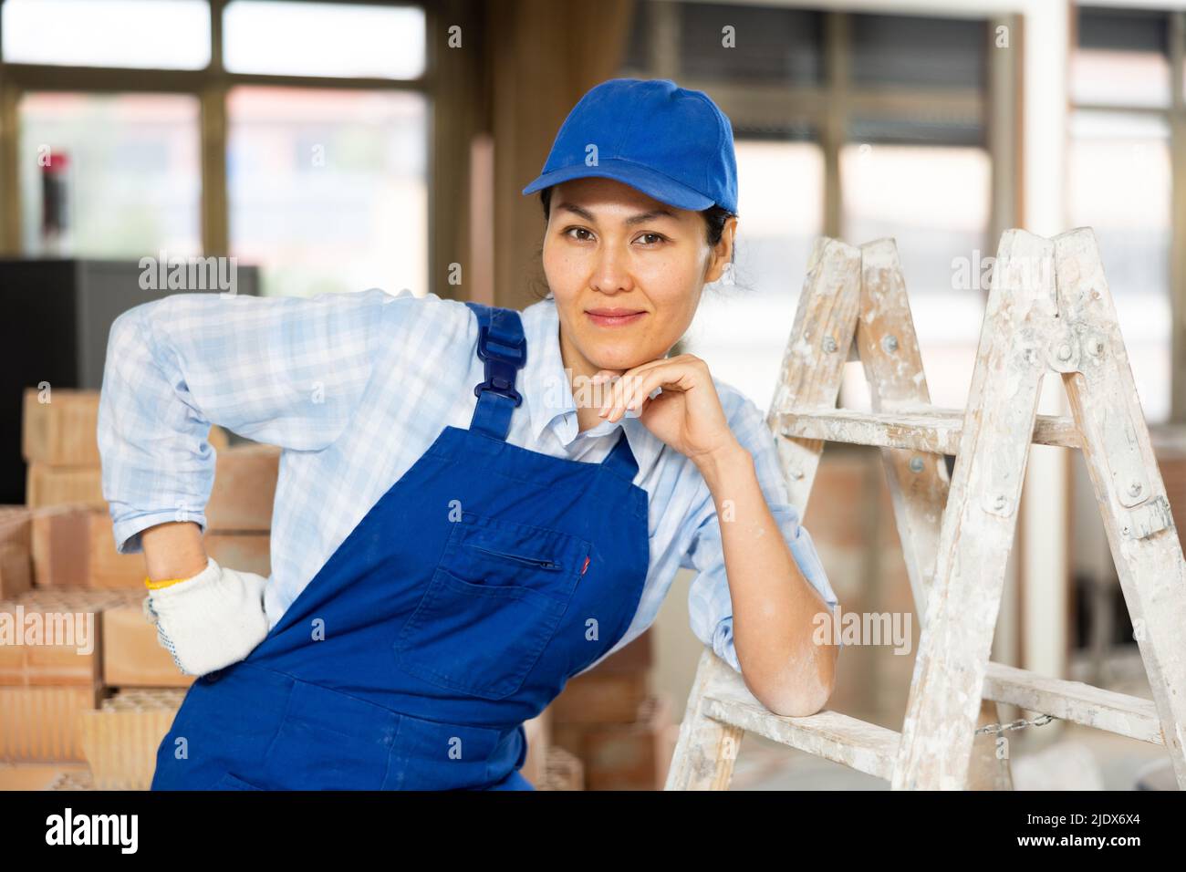 Portrait of positive builder woman in blue overalls next to stepladder ...