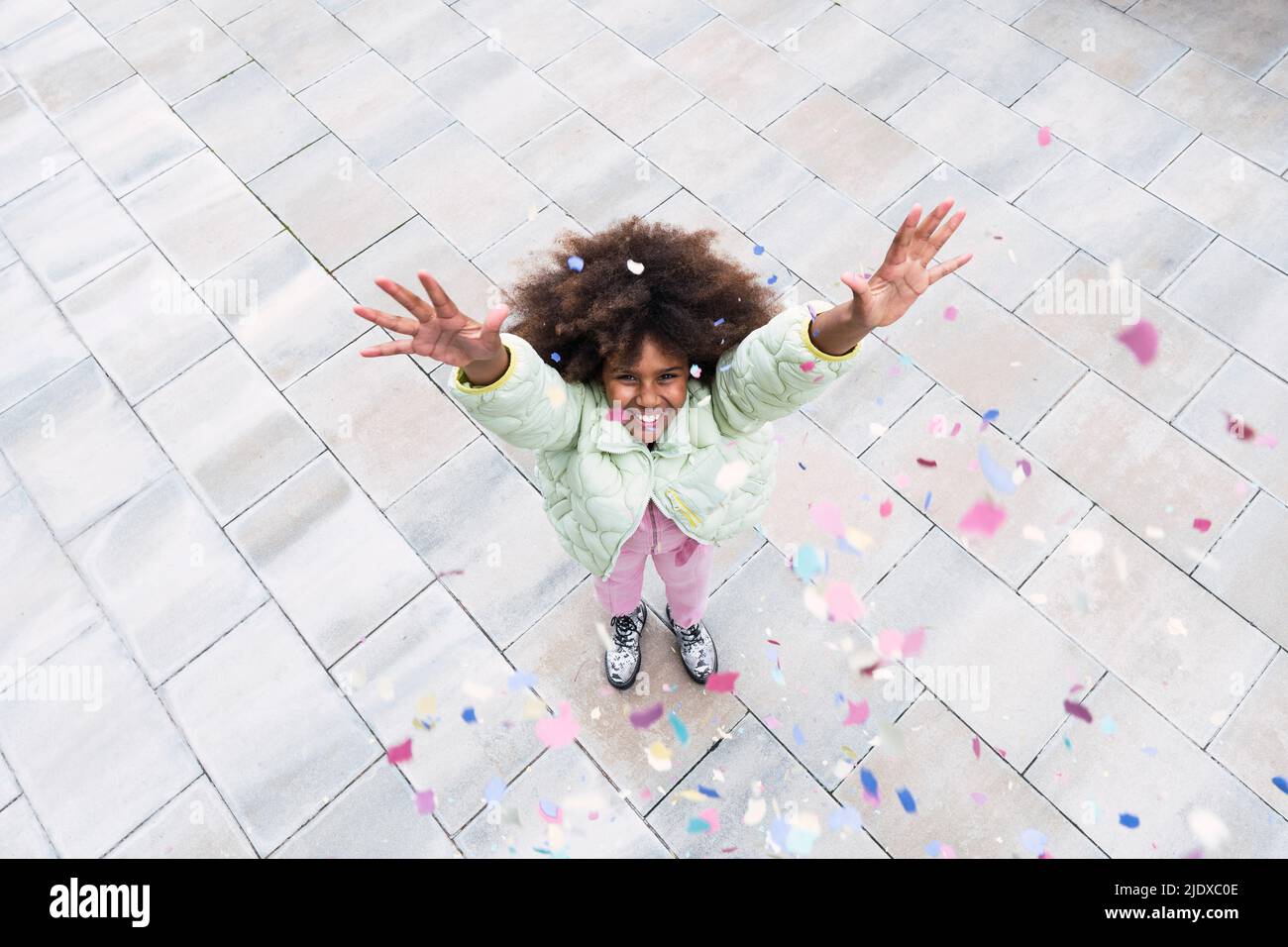 Cheerful Afro girl with arms raised standing amidst confetti falling on ...