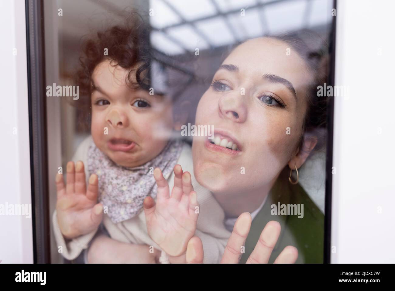 Mother and daughter looking through window pressing faces on glass pane ...