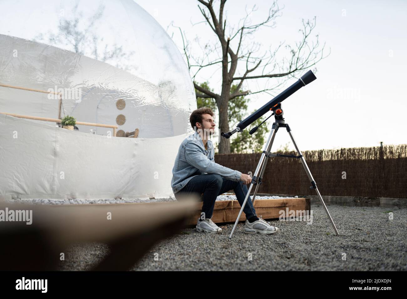 Young man with telescope sitting in front of transparent dome hotel ...