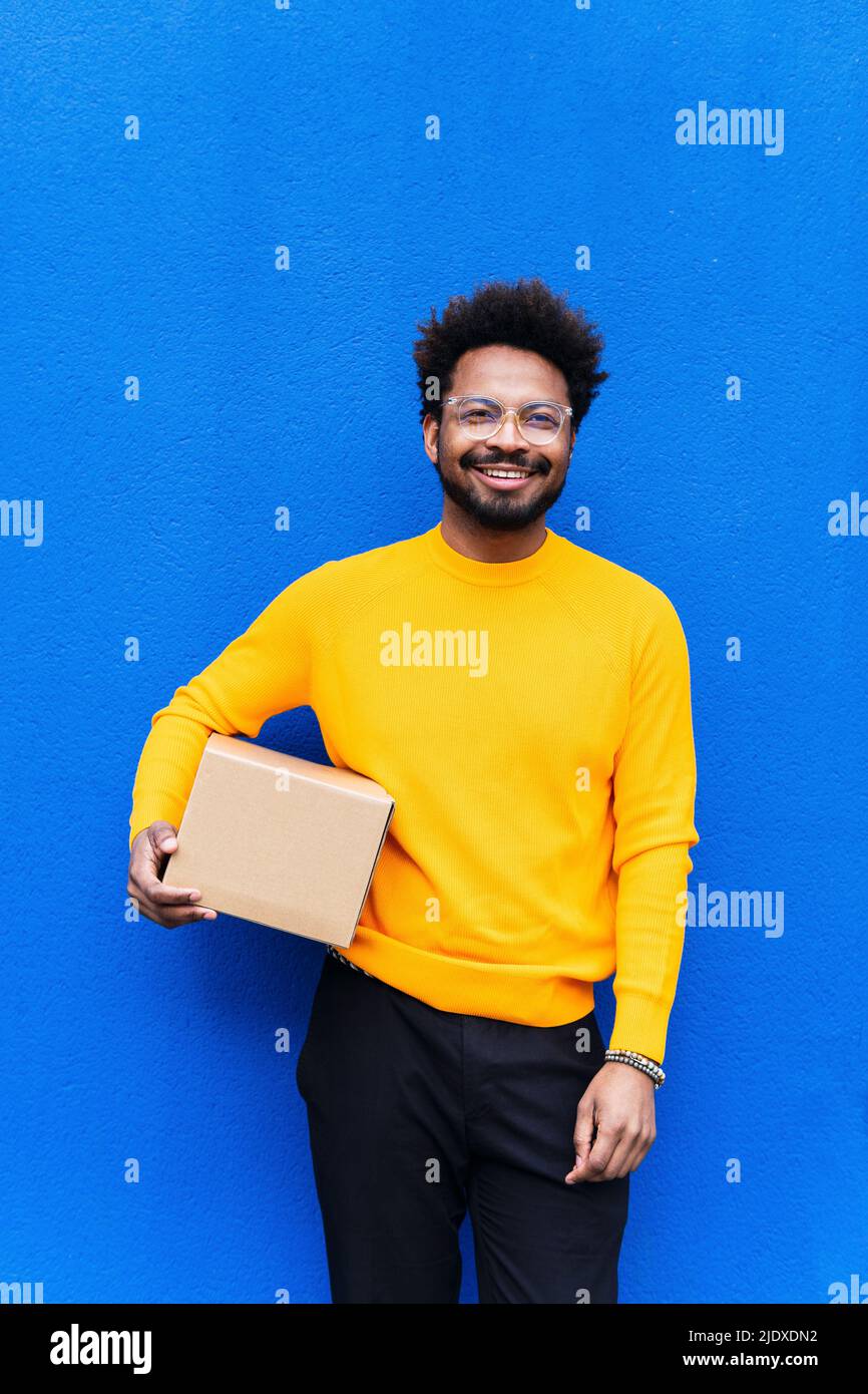 Smiling delivery man holding box standing in front of blue wall Stock ...