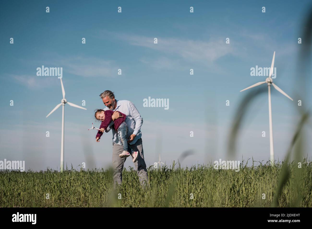 Playful father carrying daughter holding wind turbine model in field ...