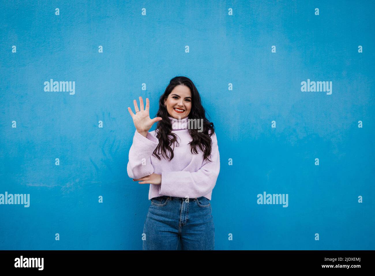 Smiling young beautiful woman showing number 5 in front of blue wall ...