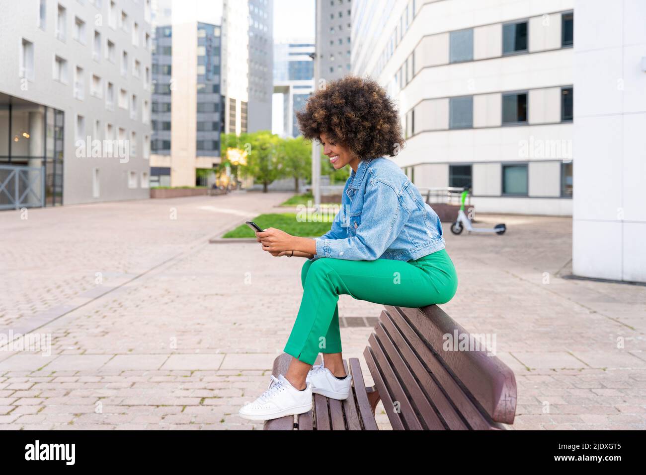 Smiling Afro woman text messaging through smart phone sitting on bench ...