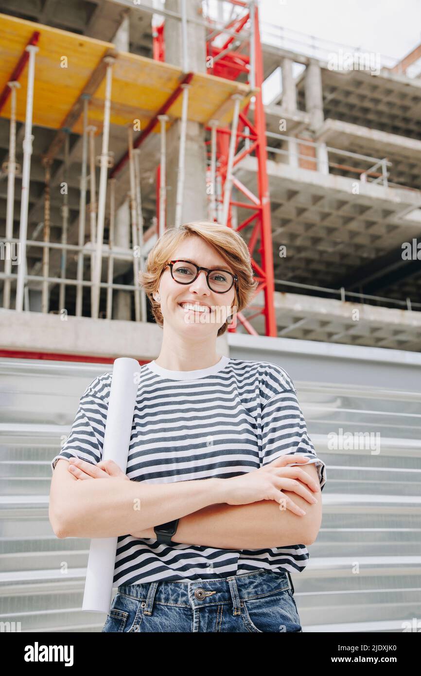 Smiling architect with blueprint standing with arms crossed at ...