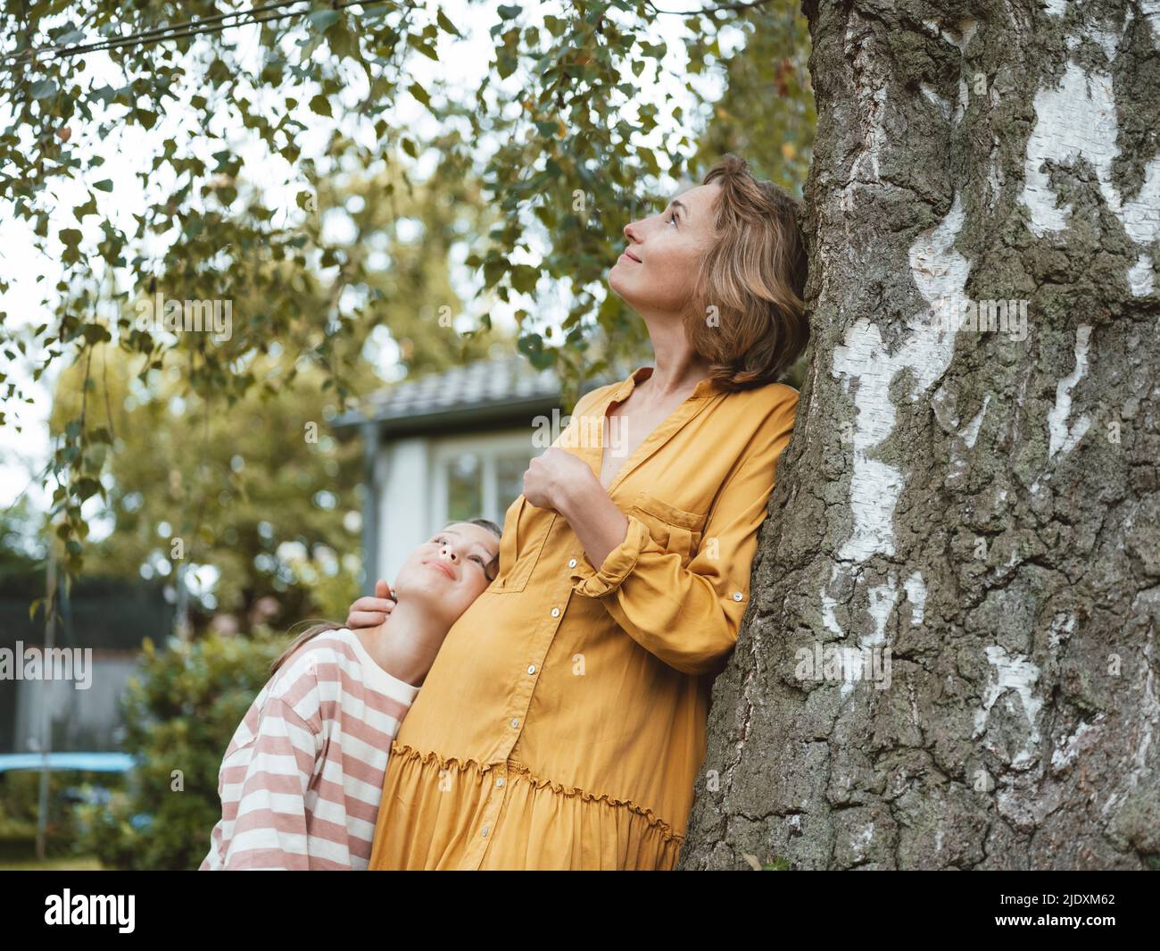 Smiling mother with daughter leaning on tree trunk Stock Photo - Alamy
