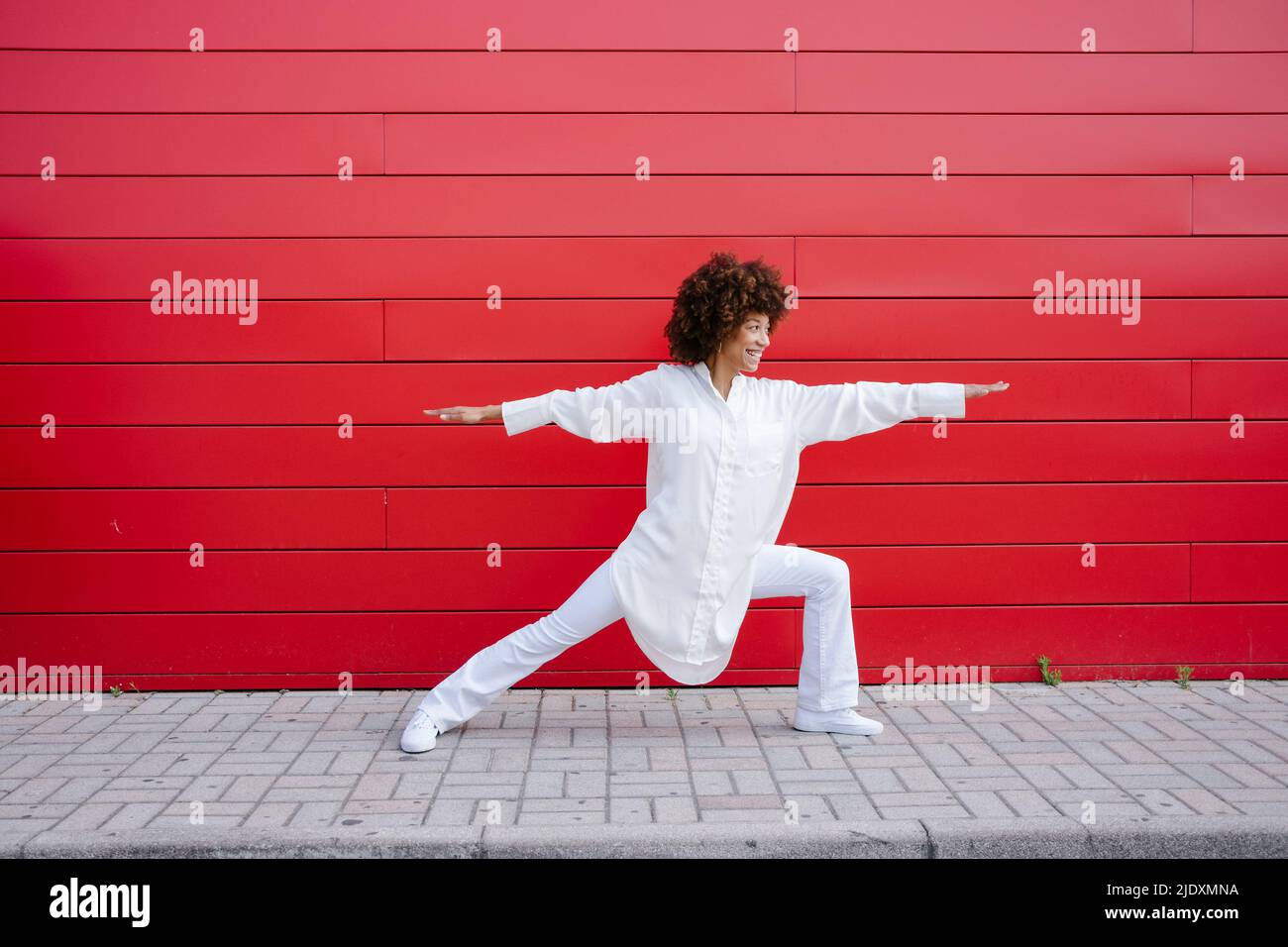 Happy woman practicing warrior position on footpath in front of red ...