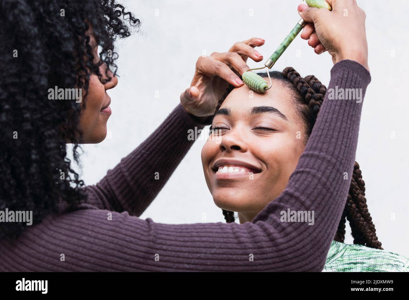 Woman Doing Facial Massage On Friend Through Jade Roller Against White