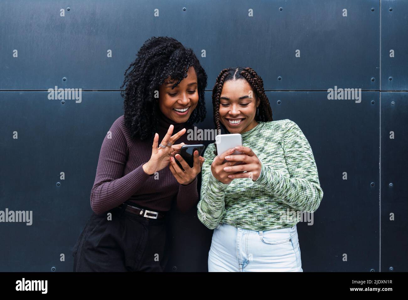 Happy young woman with friend using mobile phones in front of wall ...