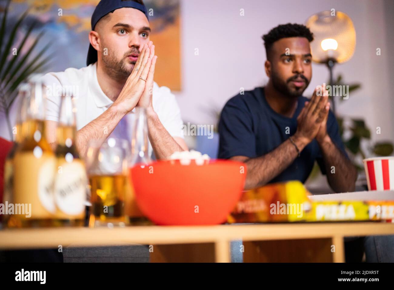 Curious young man wearing cap sitting by friend watching football match ...