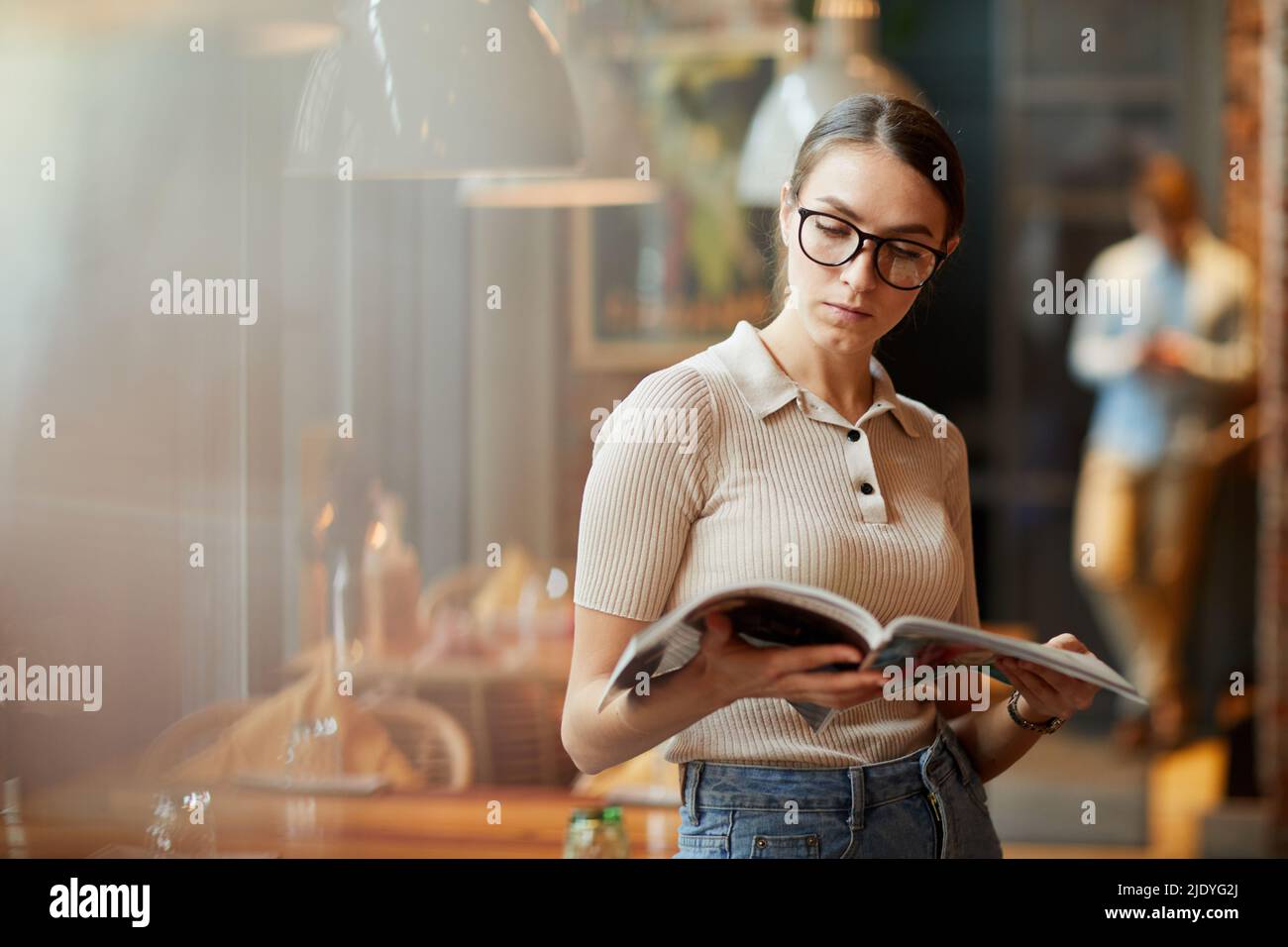 Serious intelligent young lady in glasses standing by table in cafe and ...
