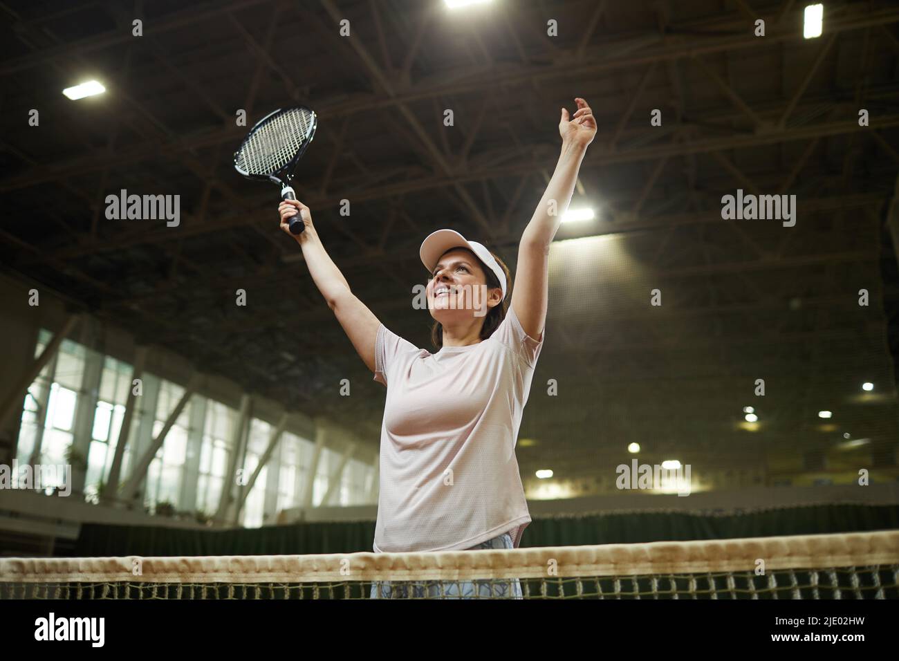 Cheerful excited young woman in cap standing by net and raising arms up ...