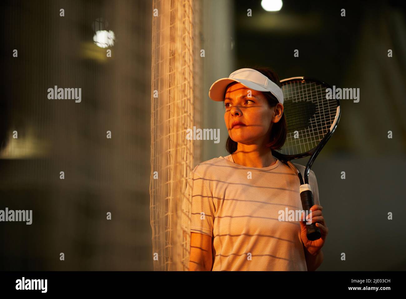 Serious brunette woman in cap standing by net and holding racket, she ...