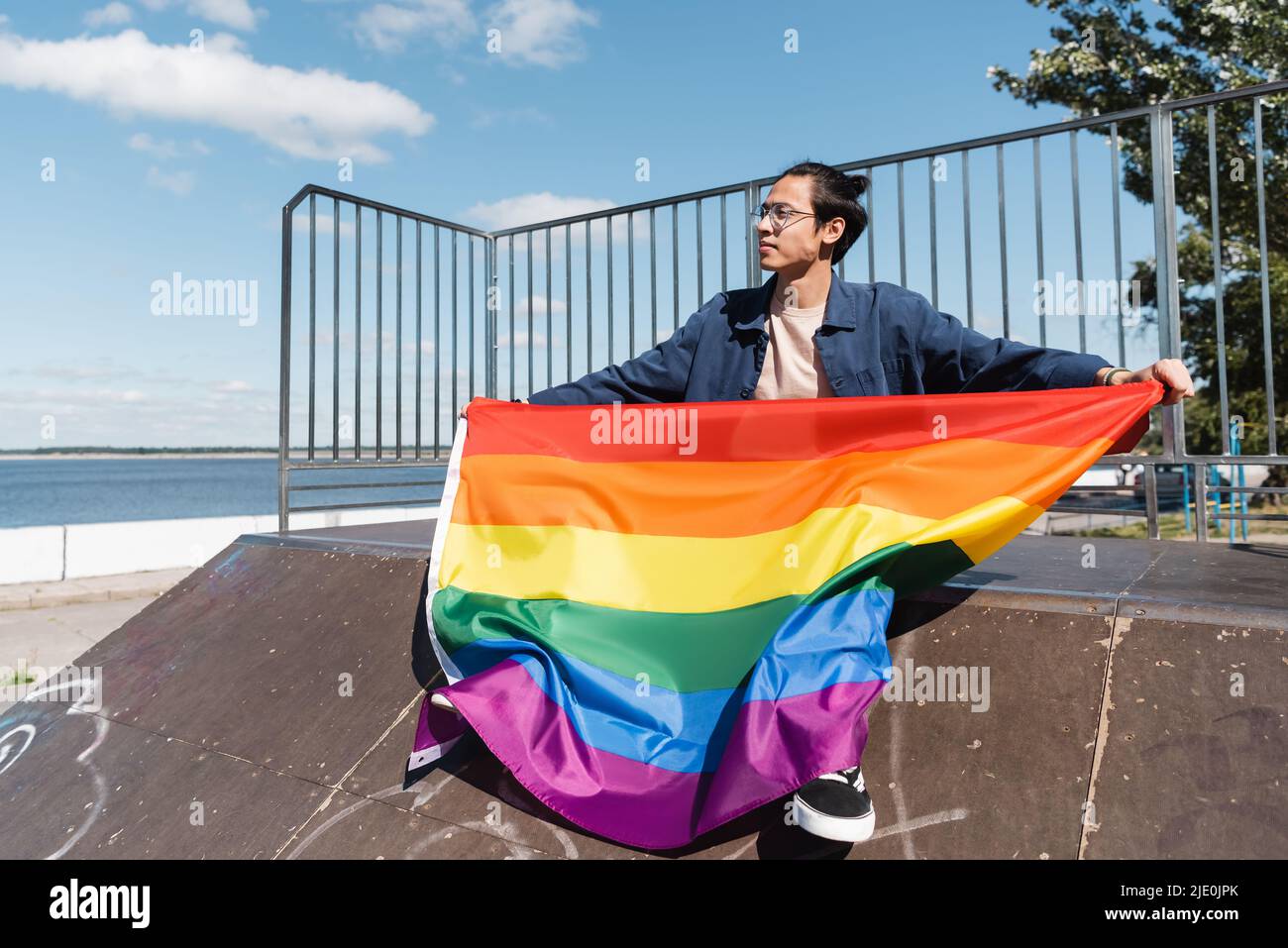 young asian man with lgbt pride flag sitting on ramp in skate park ...