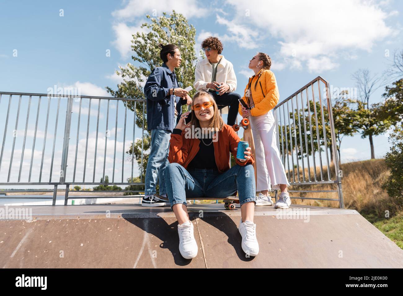 smiling woman with soda can sitting on skate ramp while multicultural ...