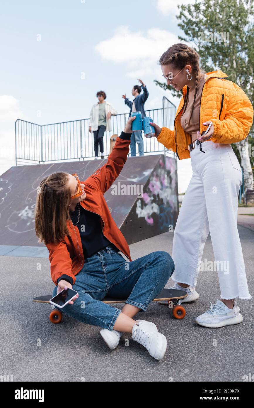 stylish women clinking soda cans near friends on skate ramp on blurred ...