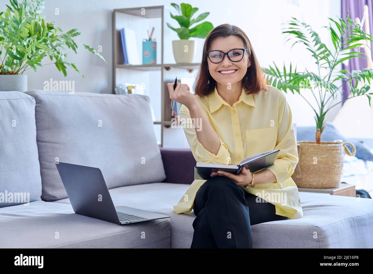 Mature business woman working on couch, using laptop looking at camera ...