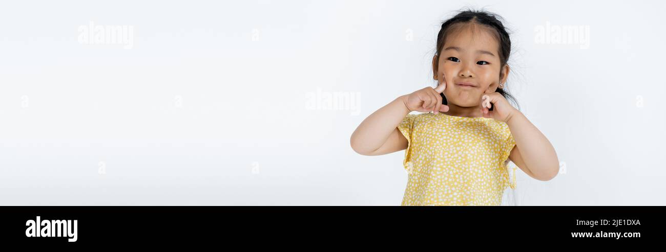 portrait of preschooler asian girl pointing at cheeks isolated on grey ...