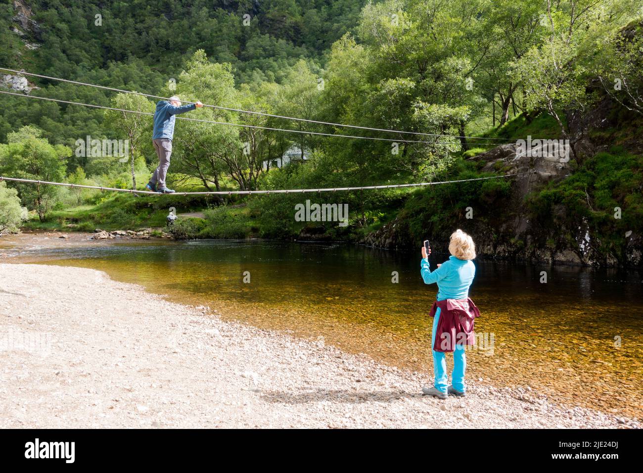 Woman photographing Man walking across Steall wire bridge over Water of ...
