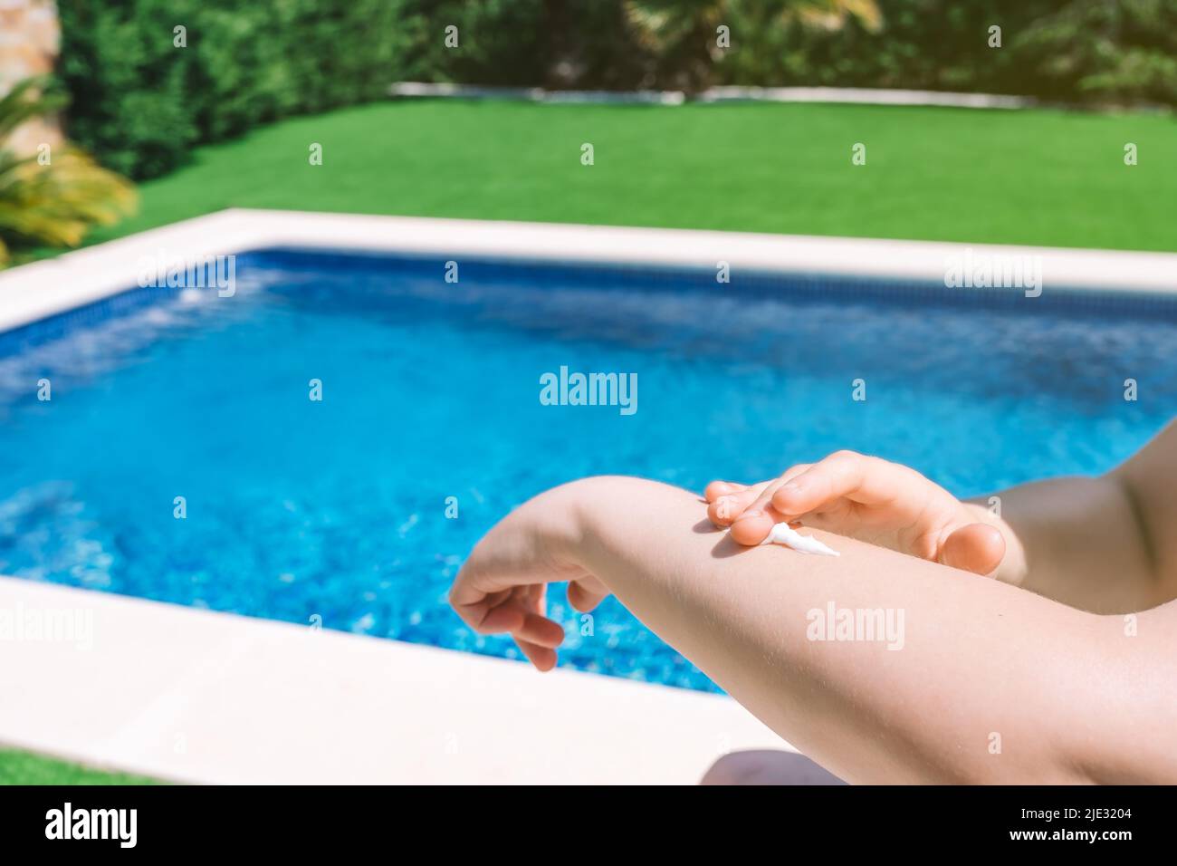 arm of a young woman applying sunscreen to her skin by a swimming pool ...